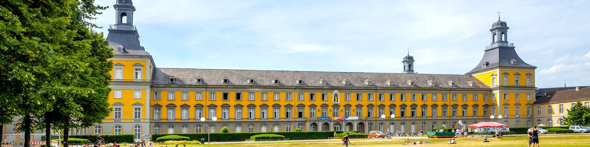 Um edifício grande e amarelo com uma torre do relógio, cercado por um pátio gramado com pessoas sentadas e caminhando.