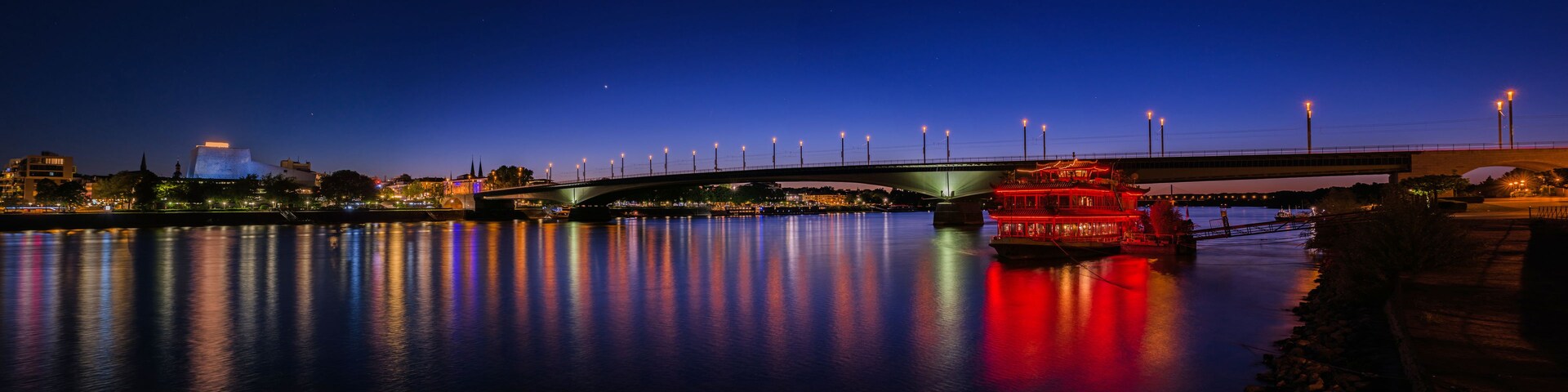 Cena noturna de um rio com uma ponte iluminada, um barco e luzes da cidade refletindo na água.