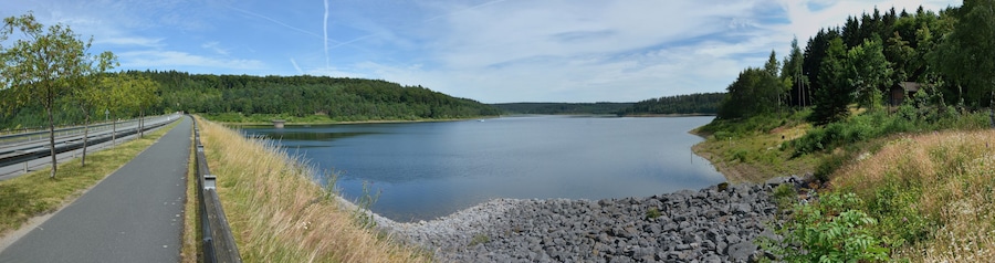Um lago tranquilo com uma alameda pavimentada à esquerda, rodeado por florestas densas e margens rochosas.