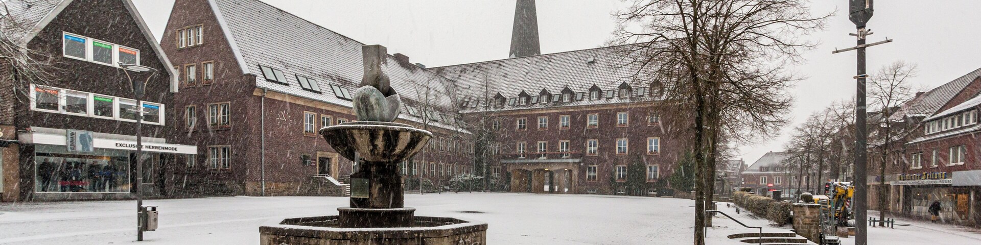 Uma praça coberta de neve, com uma fonte central, edifícios de tijolo e o topo de uma igreja ao fundo.