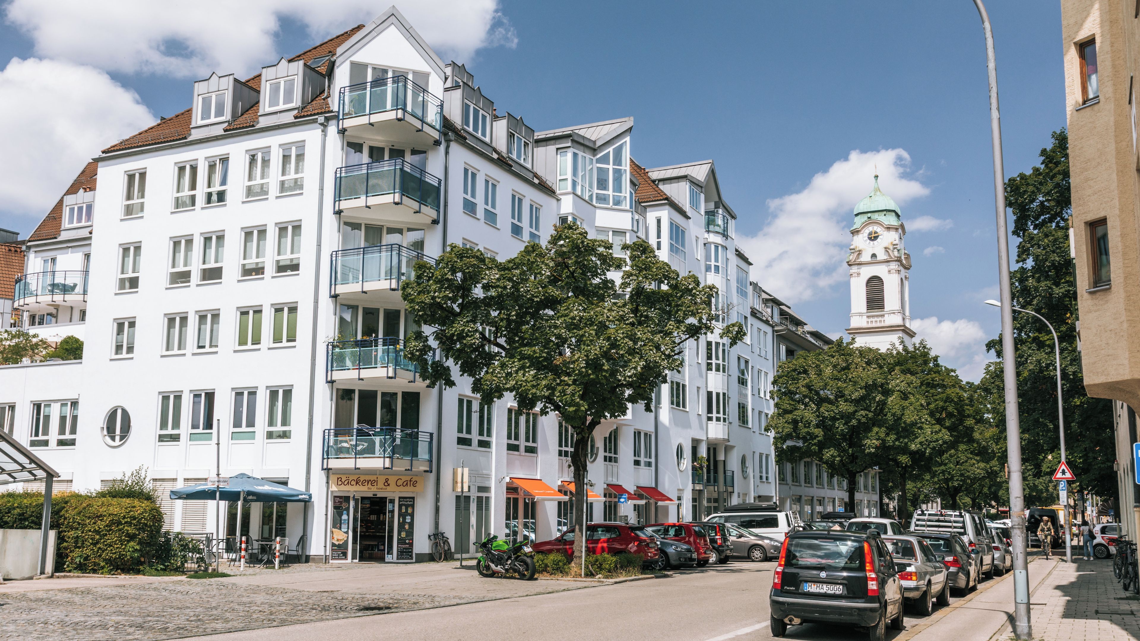 Rua com edifícios brancos, um campanário, carros estacionados e um café.