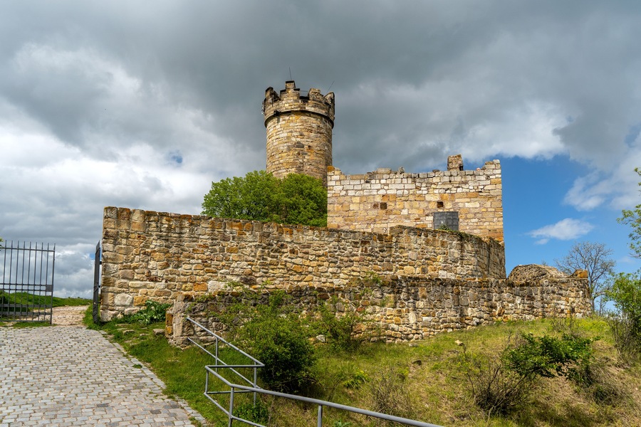 Um castelo de pedra histórico com uma torre imponente, rodeado por vegetação exuberante e um caminho de paralelepípedos.