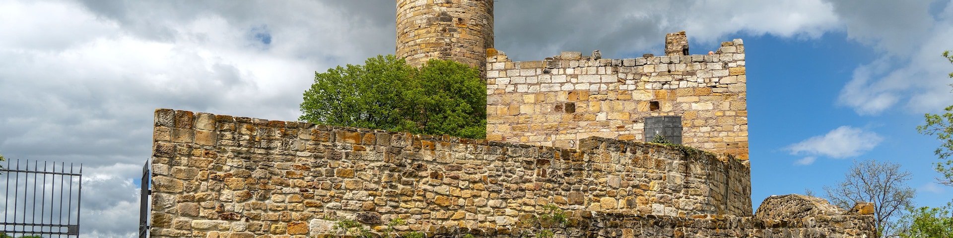 Um castelo de pedra histórico com uma torre imponente, rodeado por vegetação exuberante e um caminho de paralelepípedos.