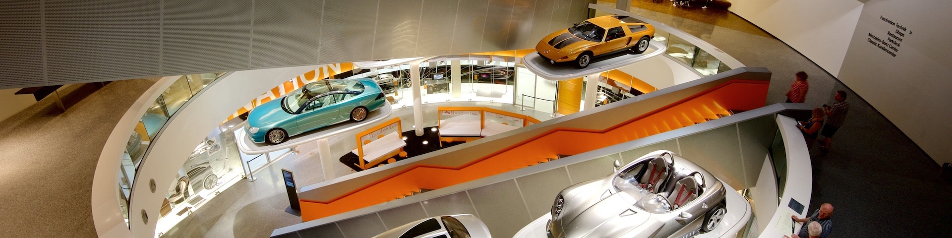 Exposição de carros em vários níveis, com diversos modelos, incluindo um carro esportivo azul e um conversível prateado.