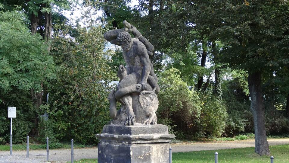 Escultura de pedra de uma figura humana em pose dinâmica, cercada por árvores e grama verdejantes.