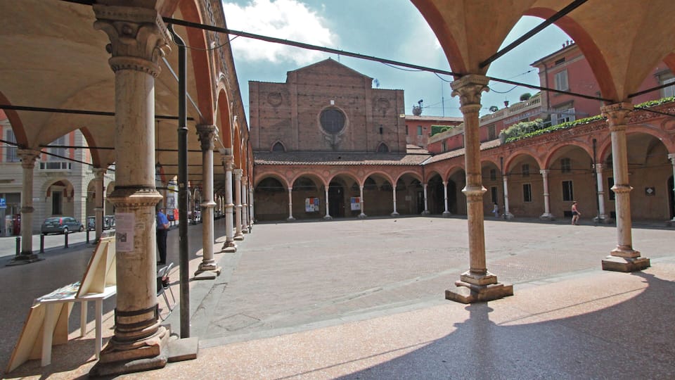 Pátio aberto com arcos e colunas, um edifício com cúpula central ao fundo, e céu claro.