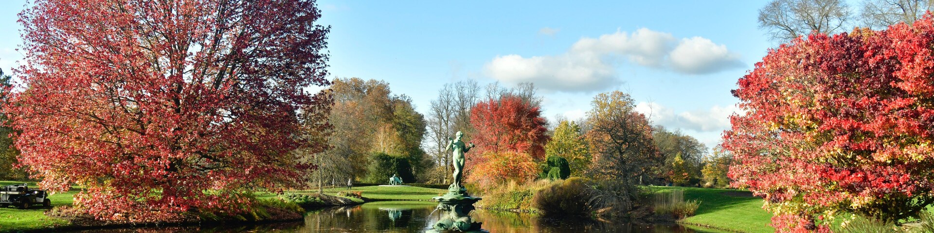Um parque tranquilo com um lago, árvores com cores de outono e uma estátua.