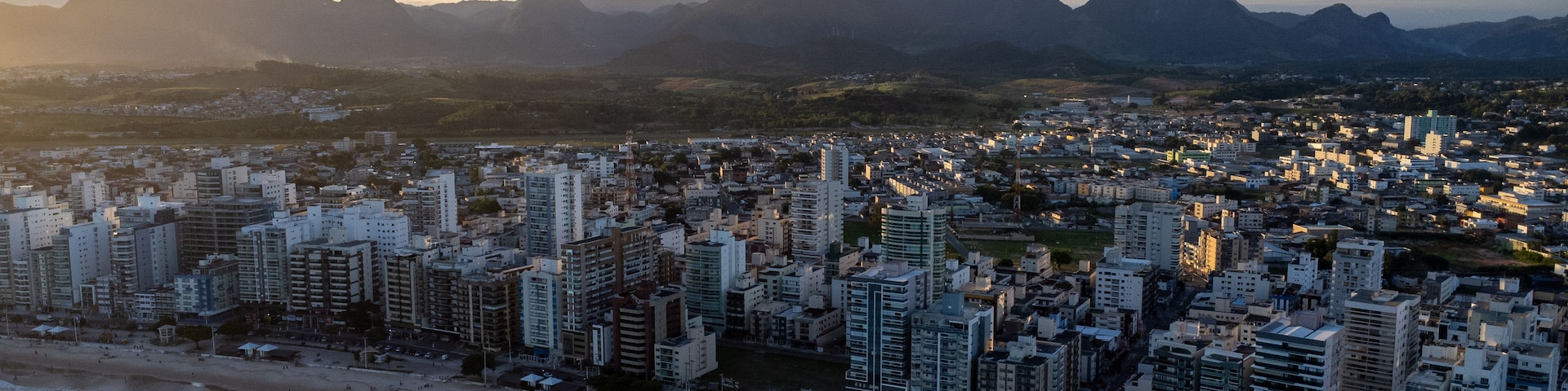 Paisagem costeira com edifícios altos, praia e montanhas ao fundo.