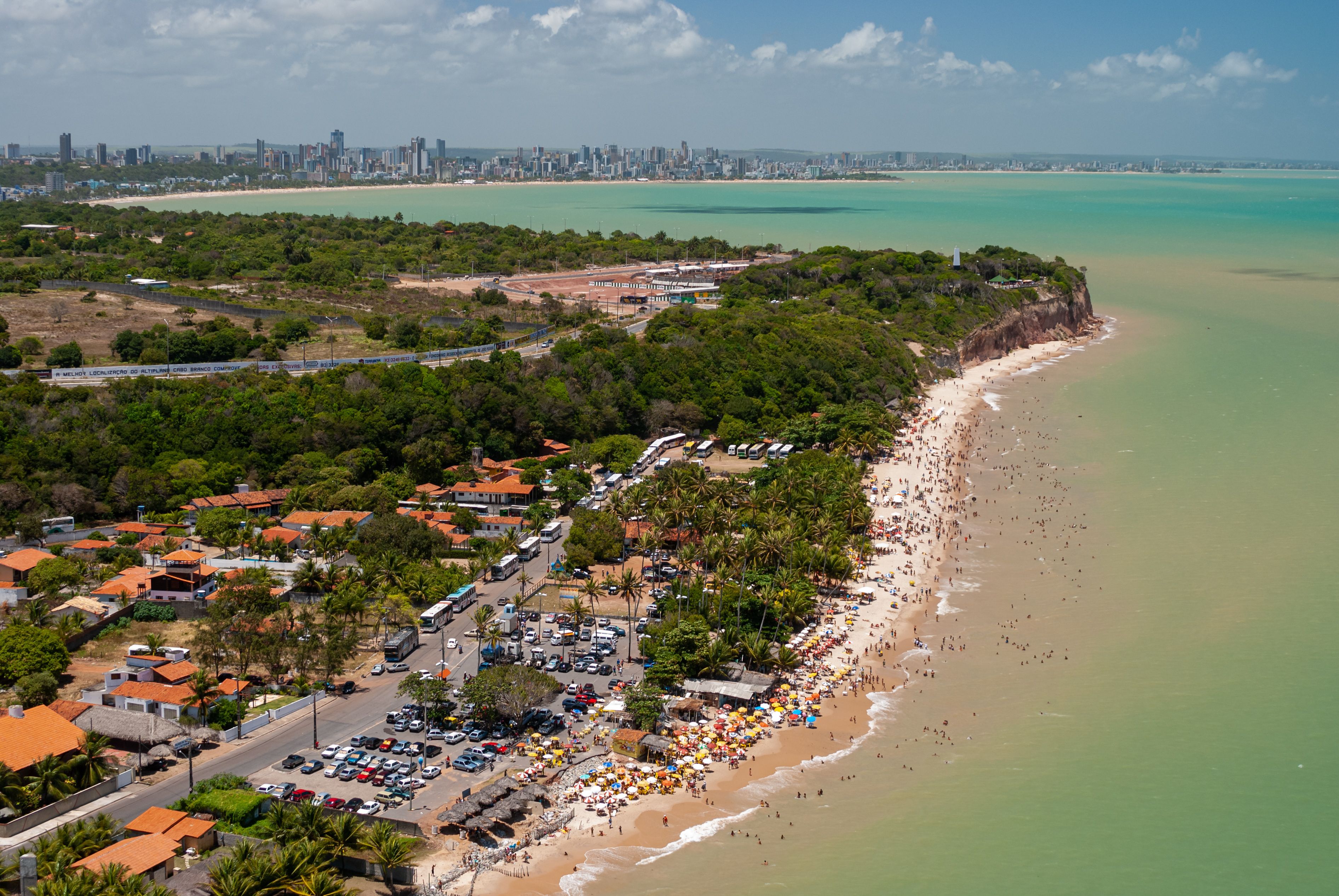 Paisagem costeira urbana com uma praia movimentada, céu limpo e horizonte urbano distante.