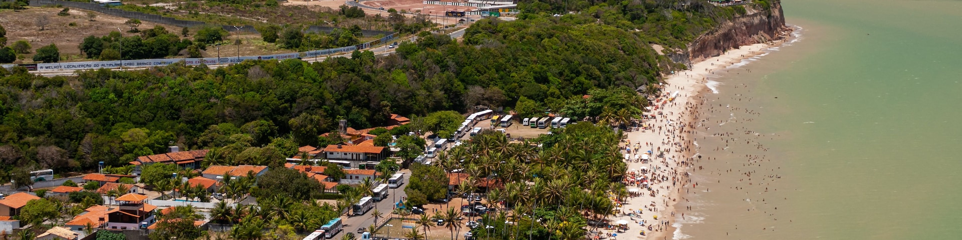 Paisagem costeira urbana com uma praia movimentada, céu limpo e horizonte urbano distante.