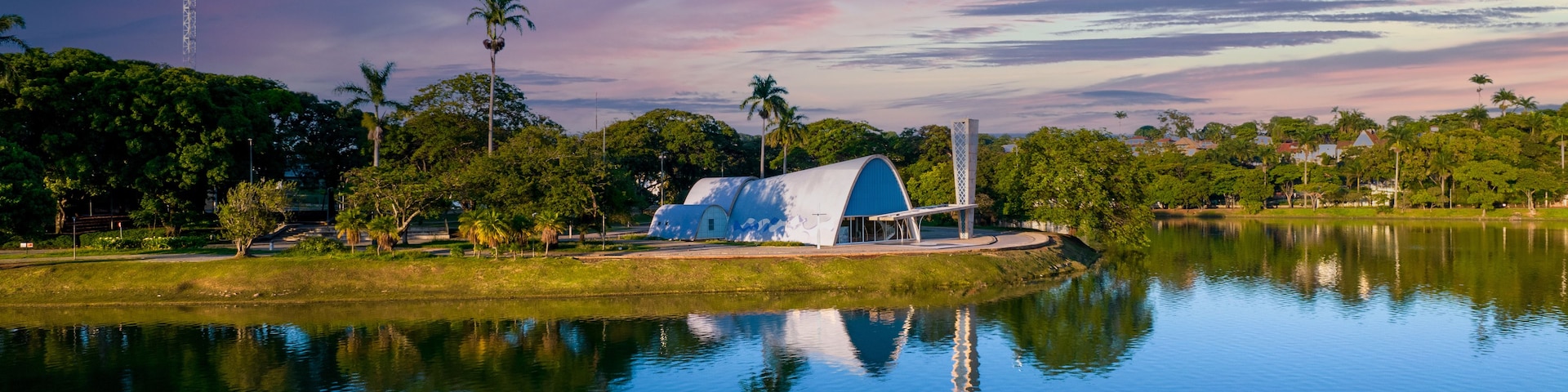 Um lago tranquilo com um edifício moderno com telhado azul, rodeado por vegetação exuberante e palmeiras.