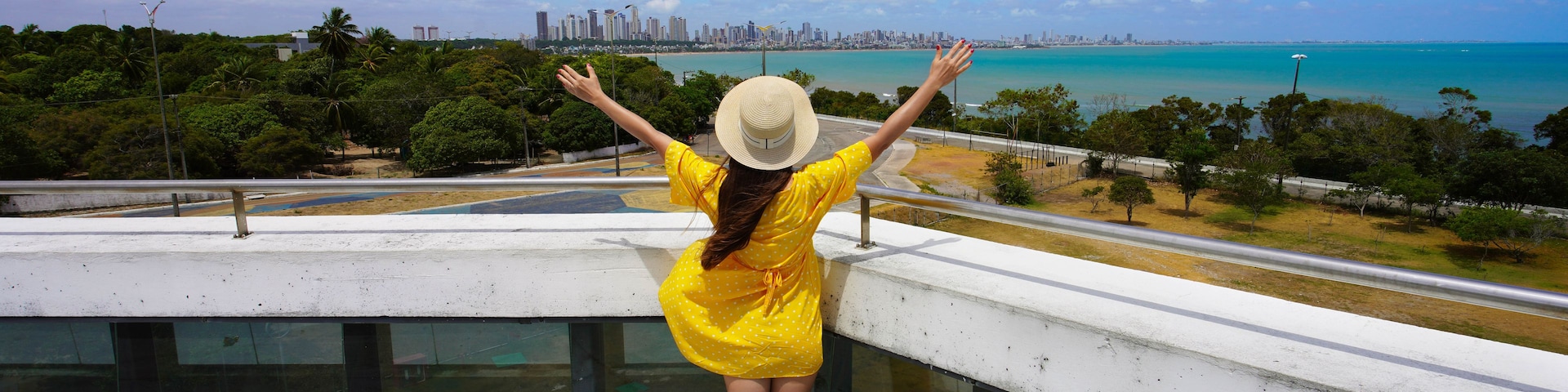 Uma pessoa num vestido amarelo, com os braços levantados, observa uma paisagem urbana costeira com edifícios e um céu azul límpido.