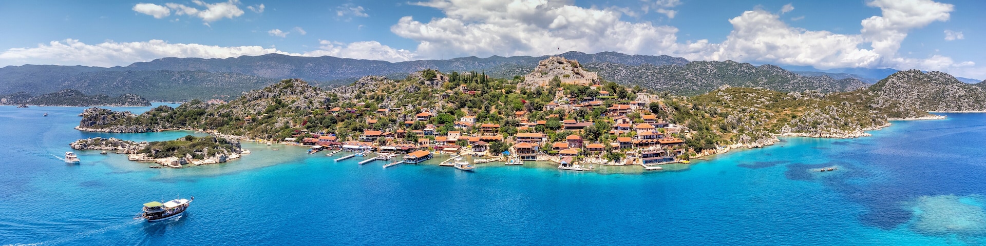 Uma vila costeira com um castelo histórico numa colina, rodeada por águas turquesa e montanhas.