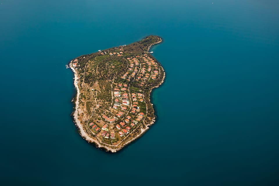 Vista aérea de uma ilha costeira com um conjunto denso de edifícios com telhados vermelhos, rodeada por uma praia arenosa estreita e um mar azul profundo.