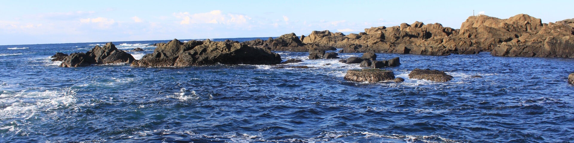 Litoral rochoso com ondas quebrando contra as rochas sob um céu claro.