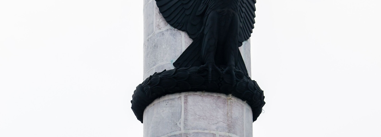 Uma estátua de uma figura alada no topo de uma coluna, com uma figura sentada abaixo.