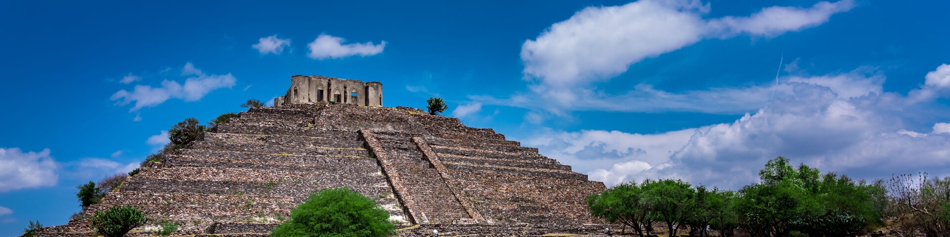 Uma pirâmide grande com uma estrutura menor no topo, rodeada por árvores e um céu azul límpido.
