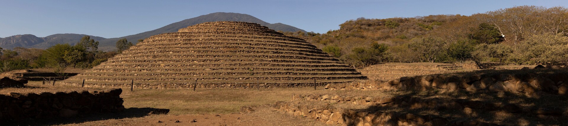 Uma grande estrutura de pedra com design escalonado, cercada por pastagem seca e árvores esparsas, com montanhas ao fundo.