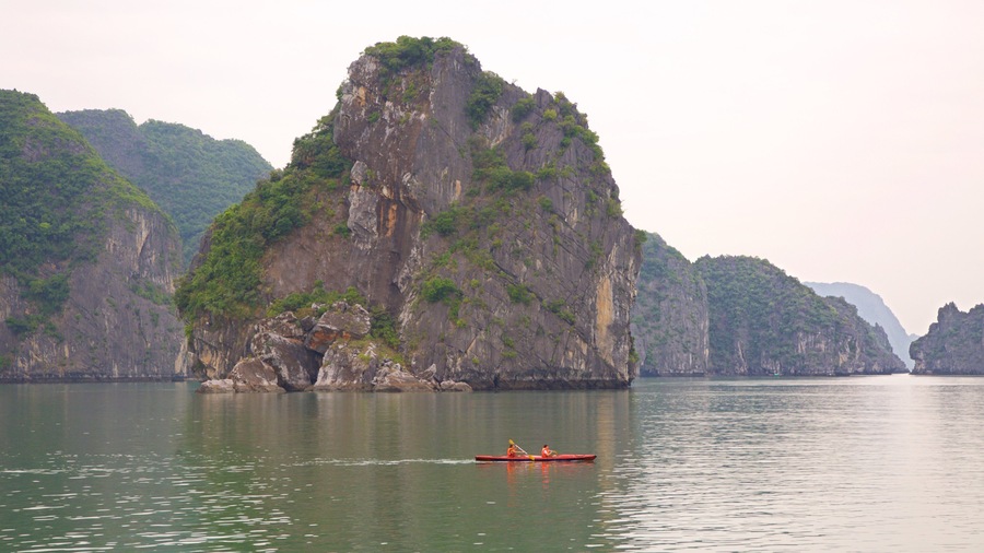 Um rio calmo com um canoa vermelha e dois remadores, cercado por colinas verdes exuberantes e um penhasco rochoso imponente.