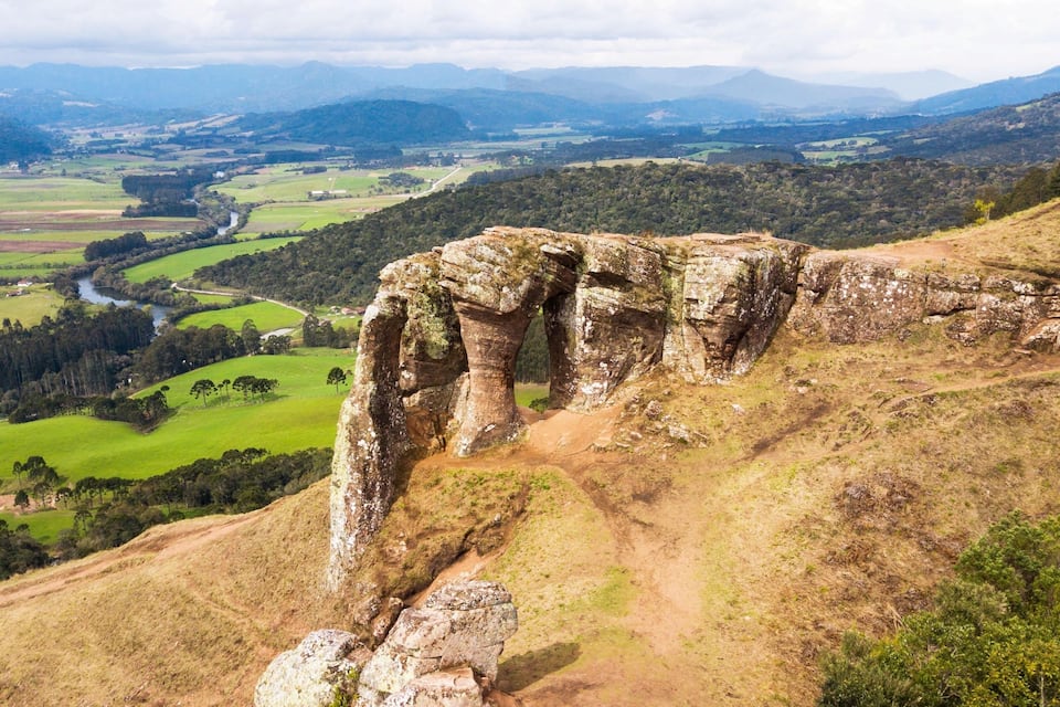 Grande formação rochosa com um arco natural, rodeada por colinas onduladas e um rio no vale abaixo.