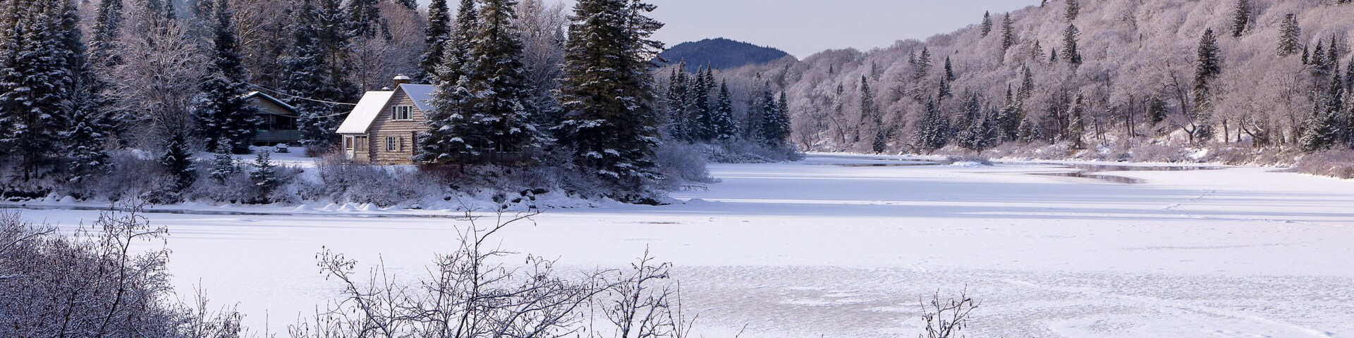 Paisagem coberta de neve com uma casa, árvores perenes densas e um lago congelado.