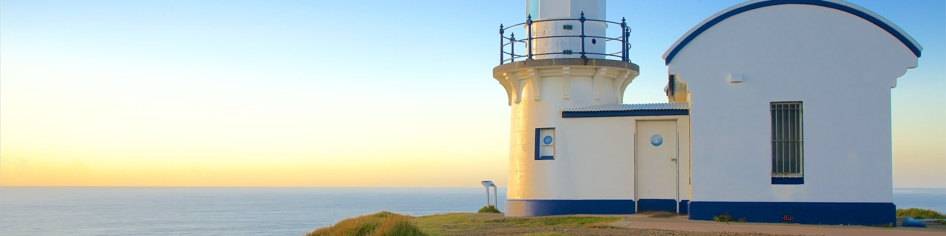Um farol com estrutura branca e azul, uma colina coberta de grama e o oceano ao fundo.