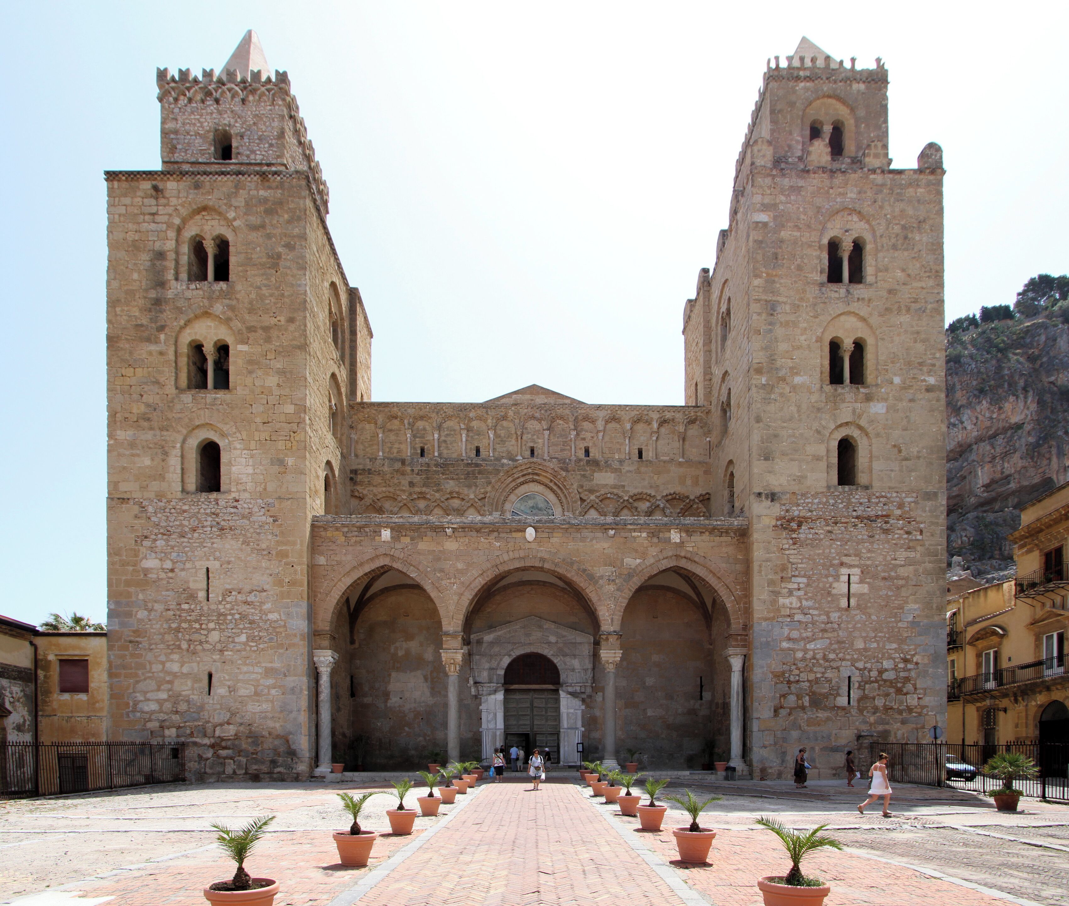 Uma catedral histórica com duas torres, um arco central e um pátio pavimentado com plantas em vasos.