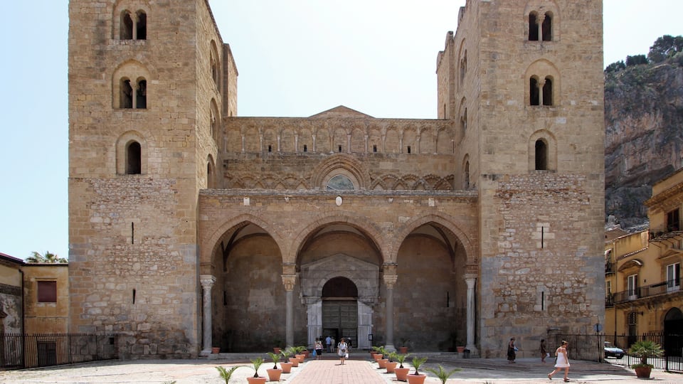 Uma catedral histórica com duas torres, um arco central e um pátio pavimentado com plantas em vasos.