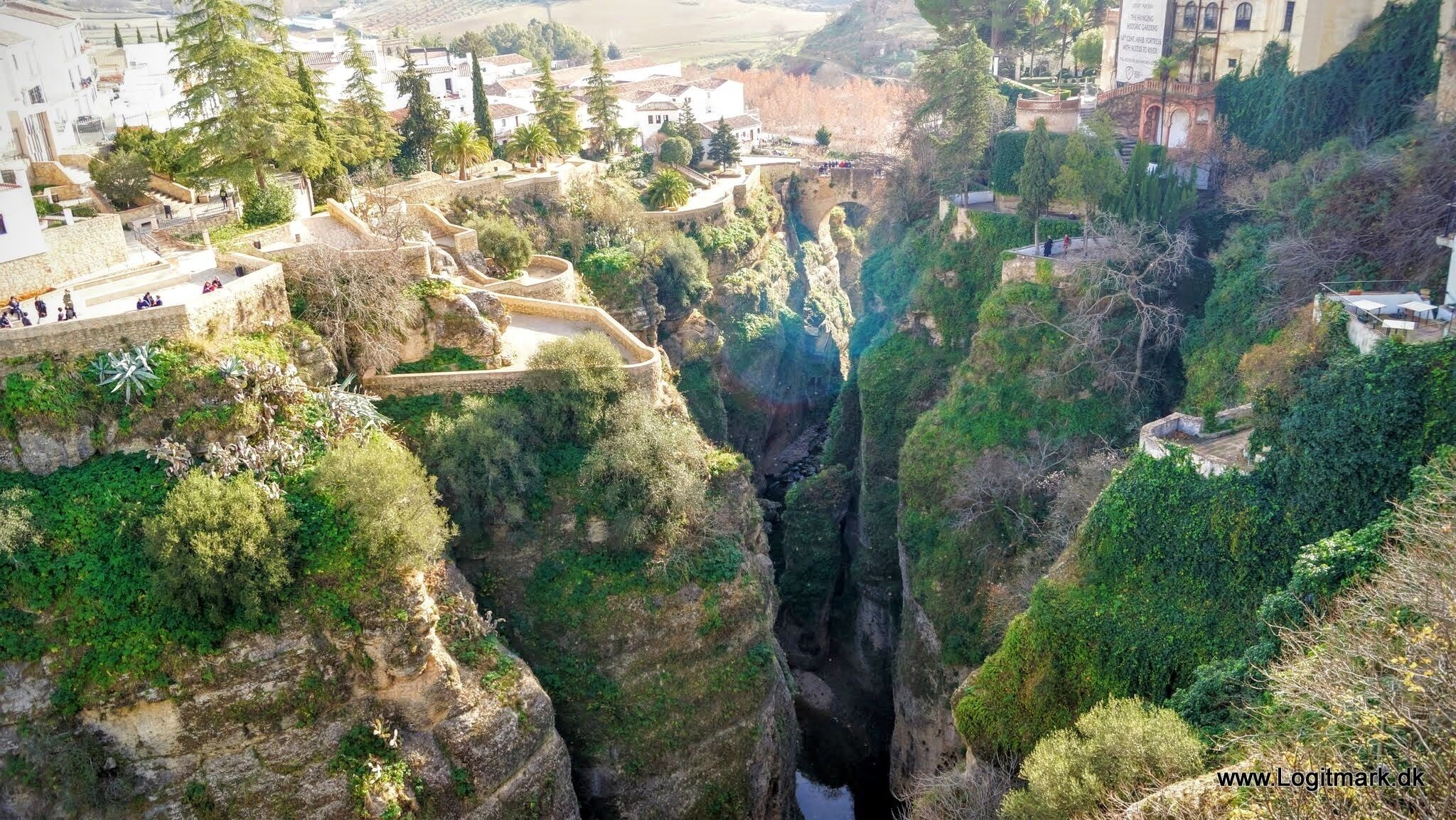Um cânion com uma ponte, vegetação exuberante e edifícios ao fundo.
