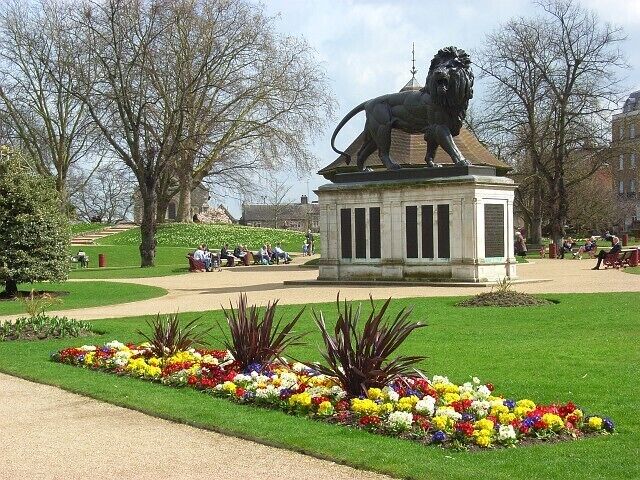 Um parque com uma estátua de leão, canteiros de flores e pessoas sentadas em bancos.