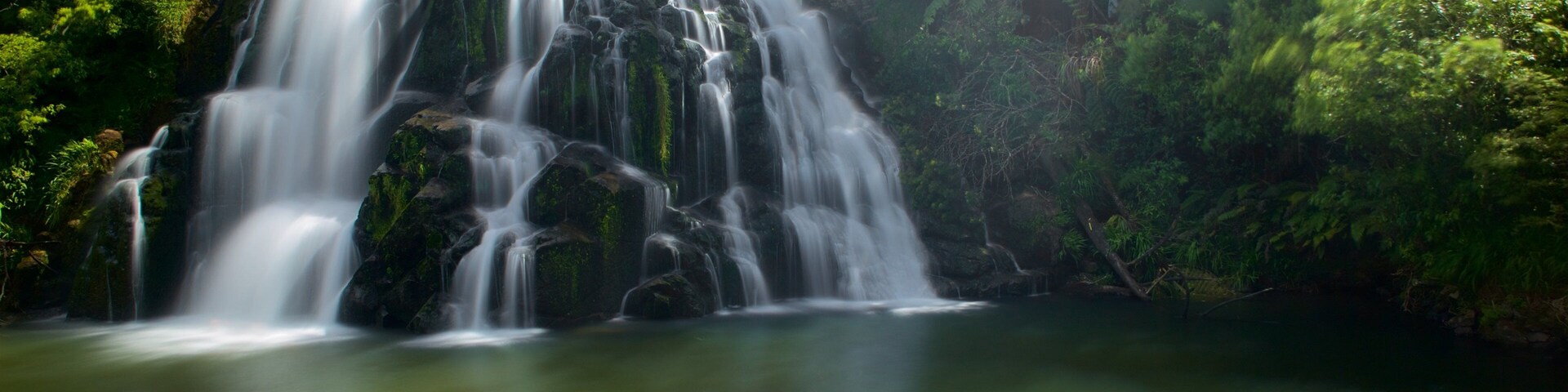 Uma cascata despencando sobre rochas em direção a um lago límpido, cercado por vegetação exuberante.