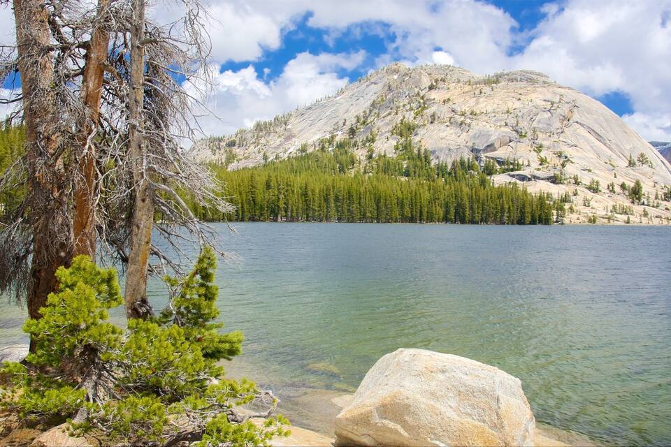 Um lago límpido cercado por montanhas, árvores e uma margem rochosa.