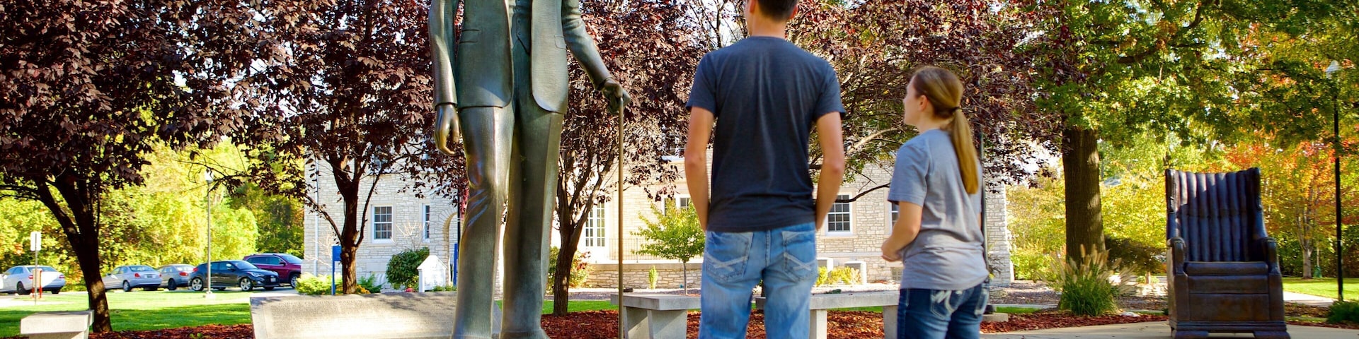 Estátua de um homem de terno em um pedestal com uma placa, cercada por árvores e um banco.