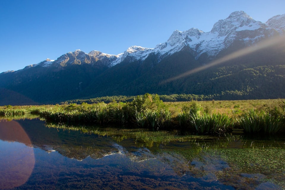Uma cordilheira com picos nevados, céu azul límpido e um corpo d'água calmo refletindo a paisagem.
