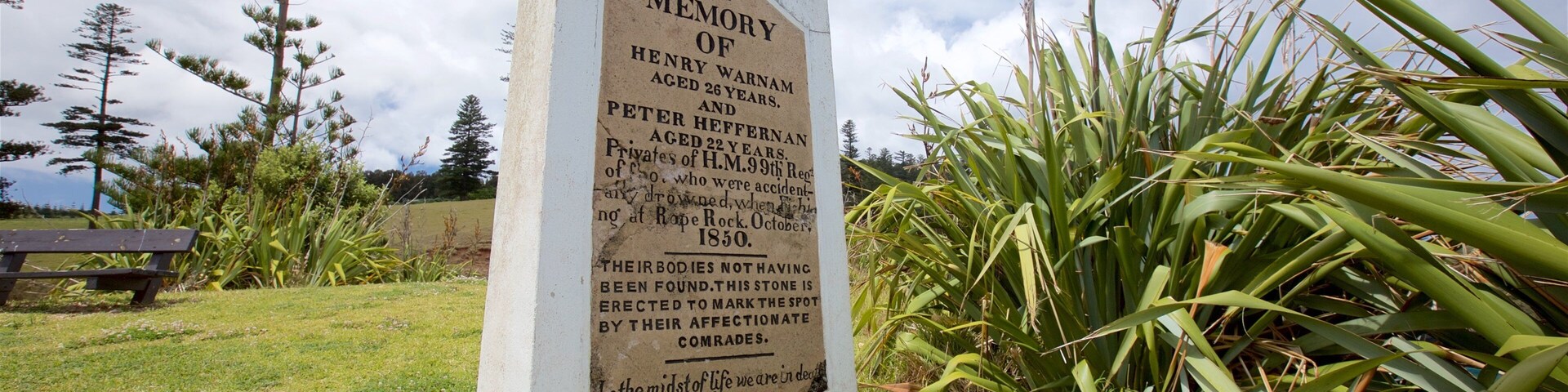 Uma pedra memorial com inscrições, cercada por grama e plantas.