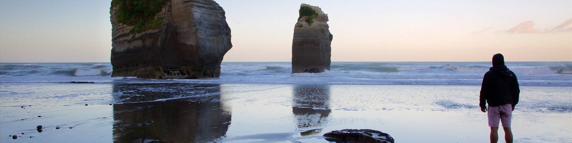 Uma pessoa em pé numa praia, com grandes formações rochosas ao fundo.