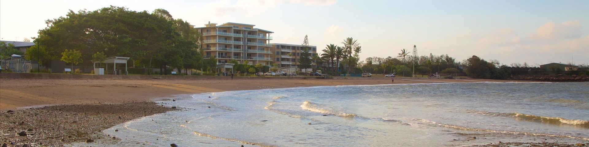 Uma praia com pedras e ondas, um edifício e árvores ao fundo.