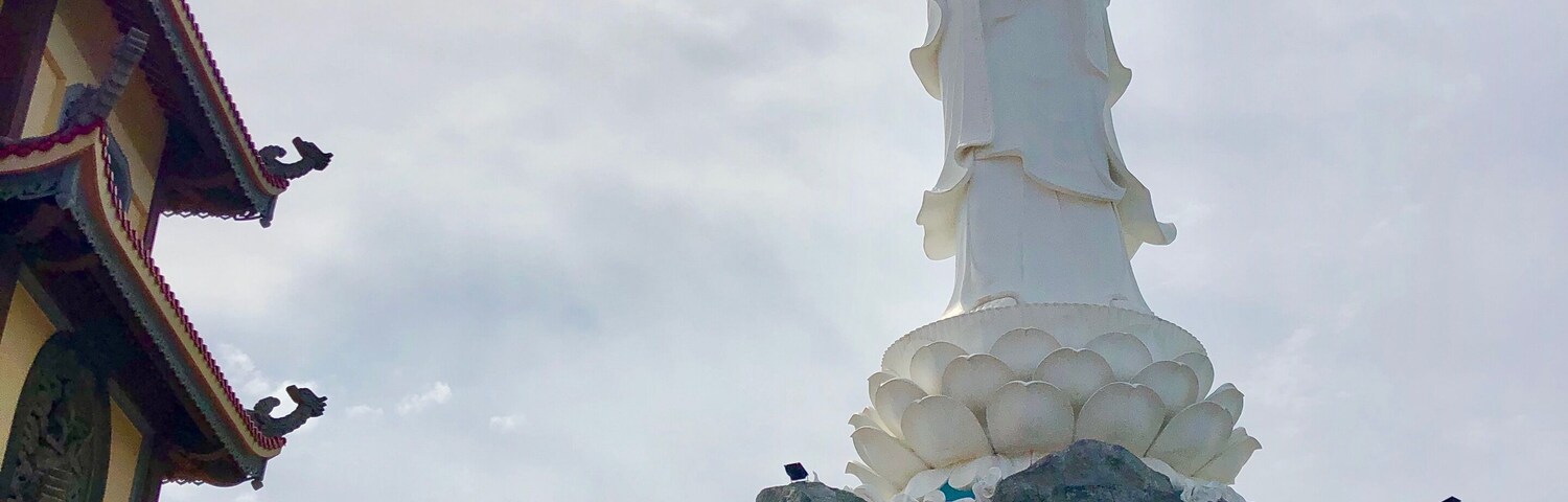 Uma grande estátua branca sobre uma rocha, com um pagode ao fundo.