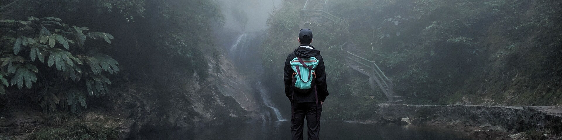 Uma pessoa em pé sobre rochas perto de uma cascata nebulosa em uma floresta.