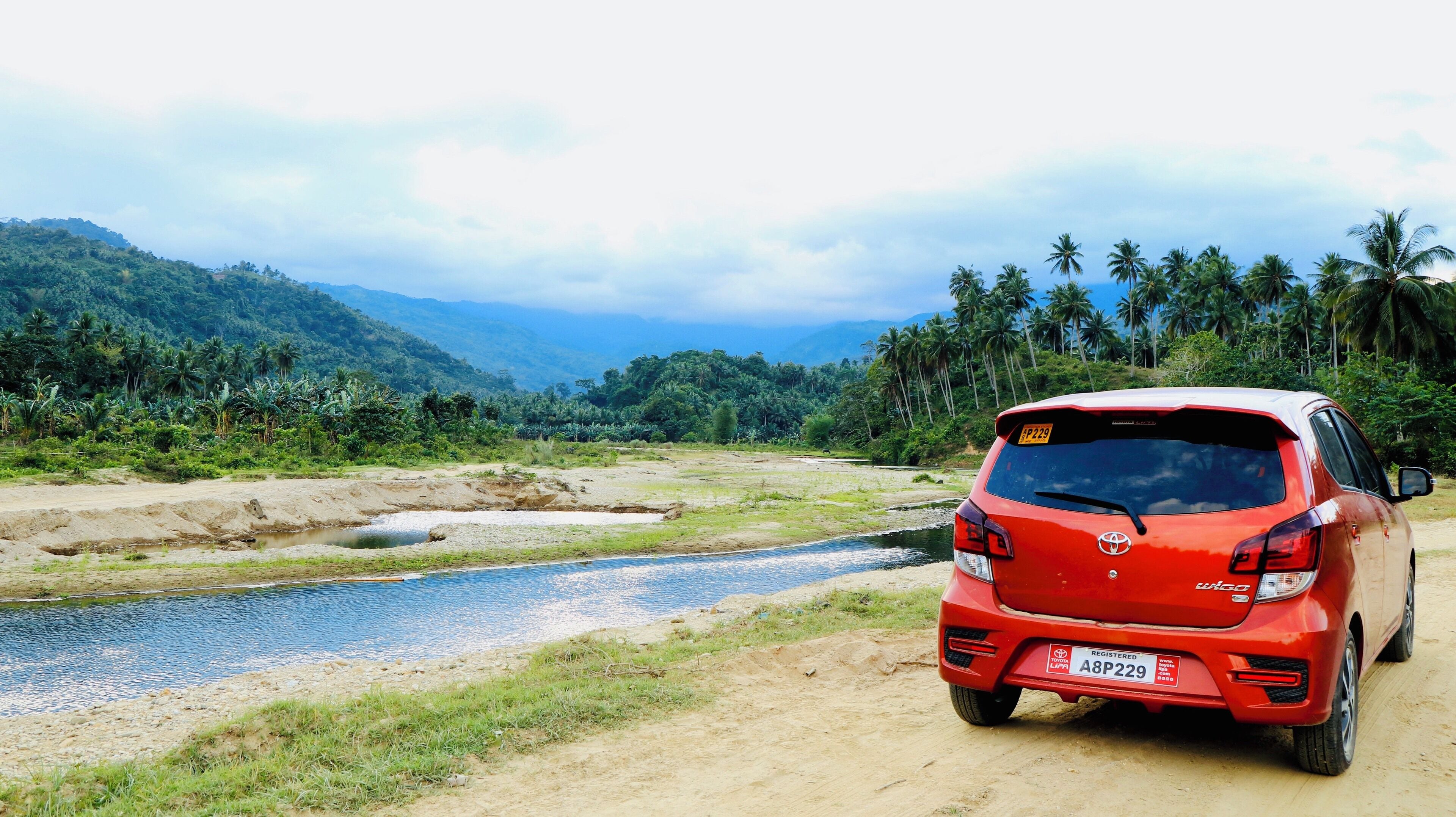 Um carro Toyota vermelho estacionado perto de um rio, com palmeiras e montanhas ao fundo.