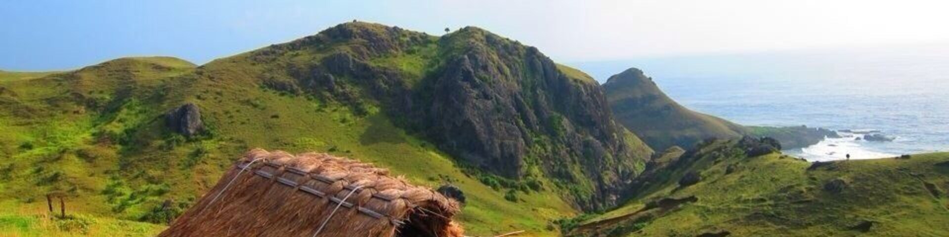 Uma cabana com telhado de palha numa encosta gramada com vista para a paisagem costeira.