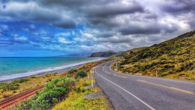 Uma estrada costeira sinuosa serpenteando por uma praia, com montanhas ao fundo e céu nublado.