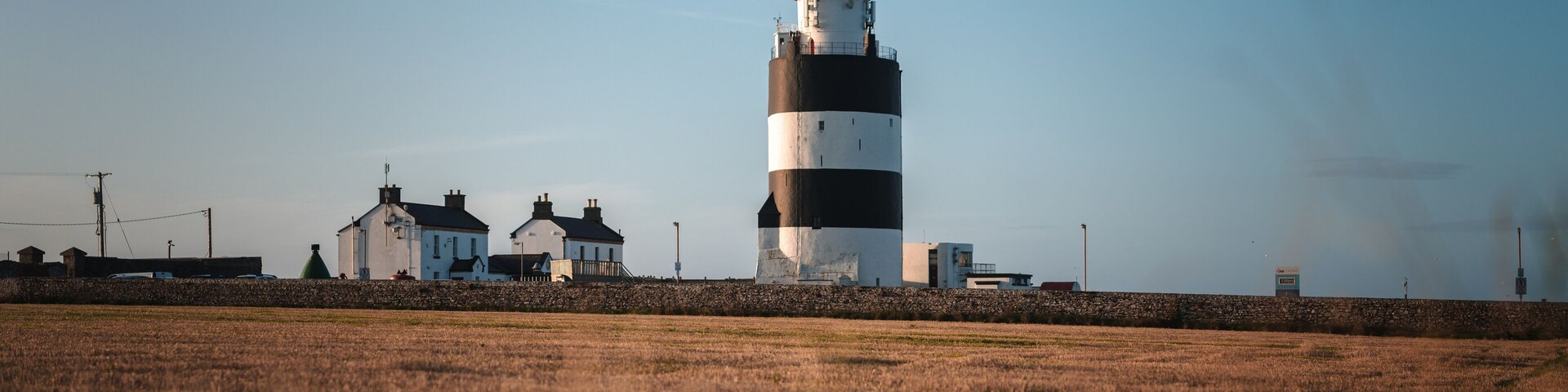 Um farol com uma torre listrada em preto e branco, cercado por um campo e algumas casas.