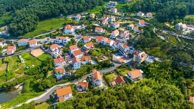 Uma aldeia densamente povoada, com casas de telhado vermelho, aninhada em um vale exuberante, cercada por colinas arborizadas.