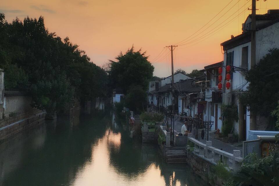 Um rio calmo ao pôr do sol, com edifícios tradicionais à direita e vegetação densa à esquerda.