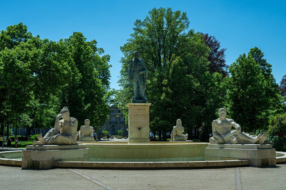 A statue of a seated man with a pedestal labeled "A.J. BRUAT" in a park with trees and a clear sky.