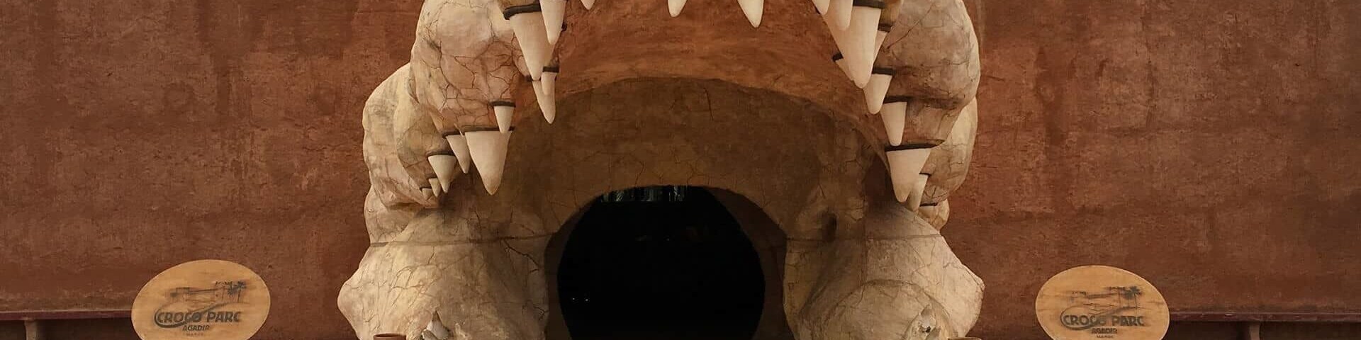 Uma grande escultura realista de boca de dinossauro, com dentes, diante de um cenário que lembra um castelo.