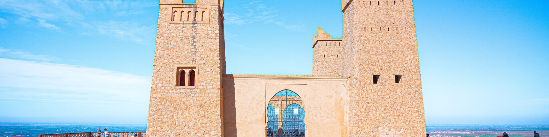 Um portão de pedra histórico com duas torres, céu azul límpido e vista para o litoral.