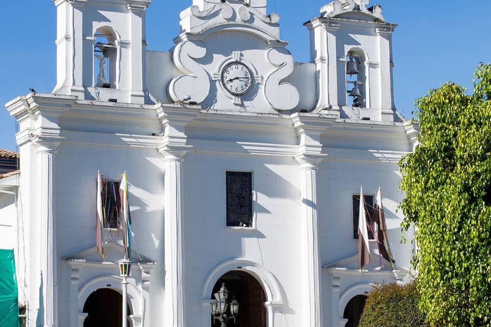 Uma igreja branca com relógio e torre do sino, rodeada por um mercado ao ar livre movimentado com toldos verdes.