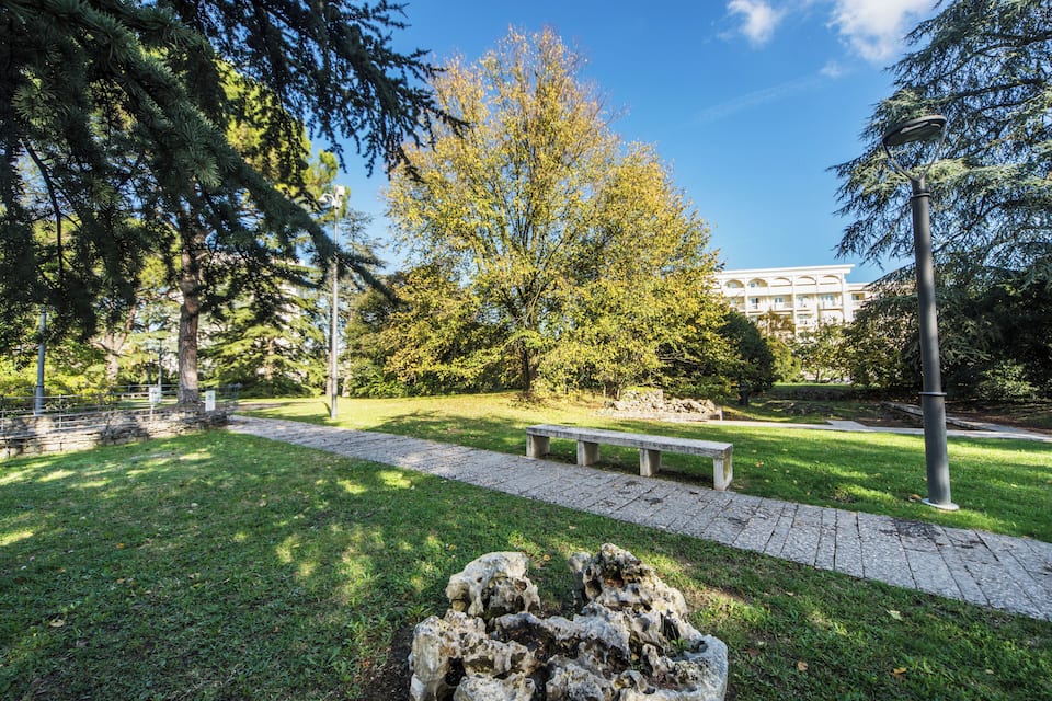 Um parque com um banco de pedra, árvores e um edifício ao fundo.